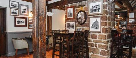 Interior rústico com parede de pedra, vigas de madeira e piso de tijolo. Quadros emoldurados adornam a parede e há um espaço de bar com garrafas.