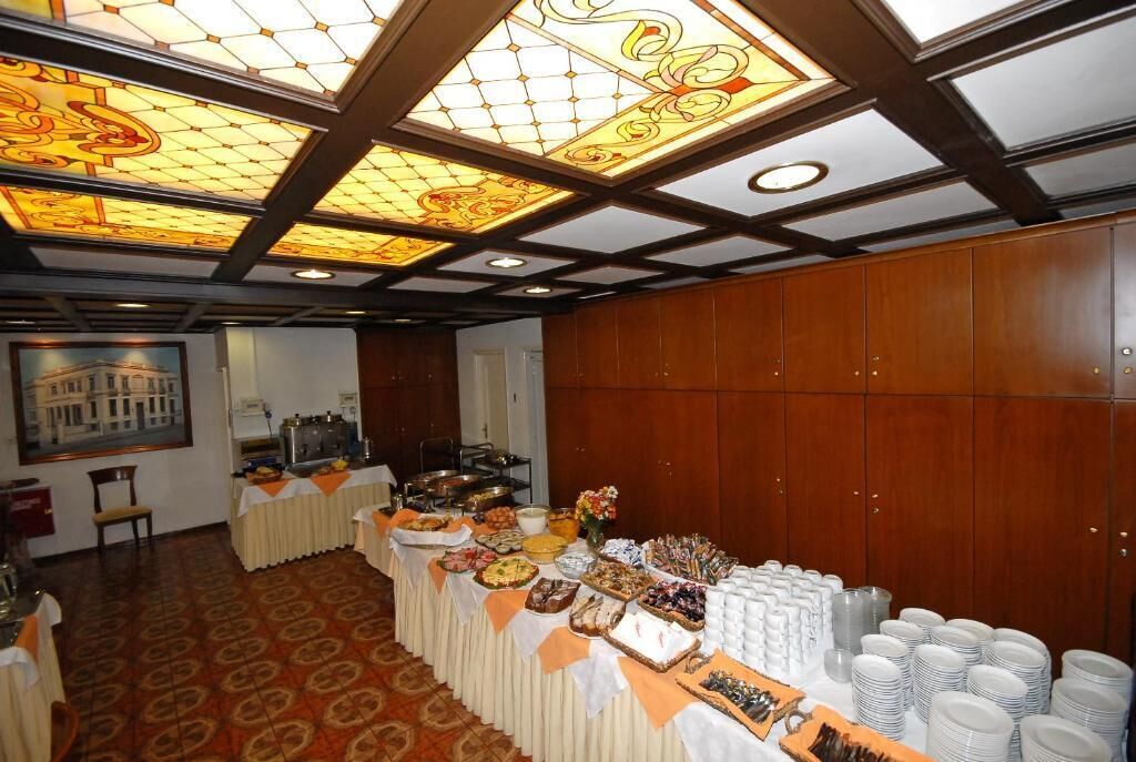 Uma sala de banquetes com uma mesa comprida preparada para um buffet, painéis de madeira nas paredes e teto com janelas de vitrais.