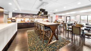 Uma área de refeições de hotel moderna, com uma longa mesa comunitária, várias cadeiras, uma cozinha ao fundo e um tapete estampado no chão.
