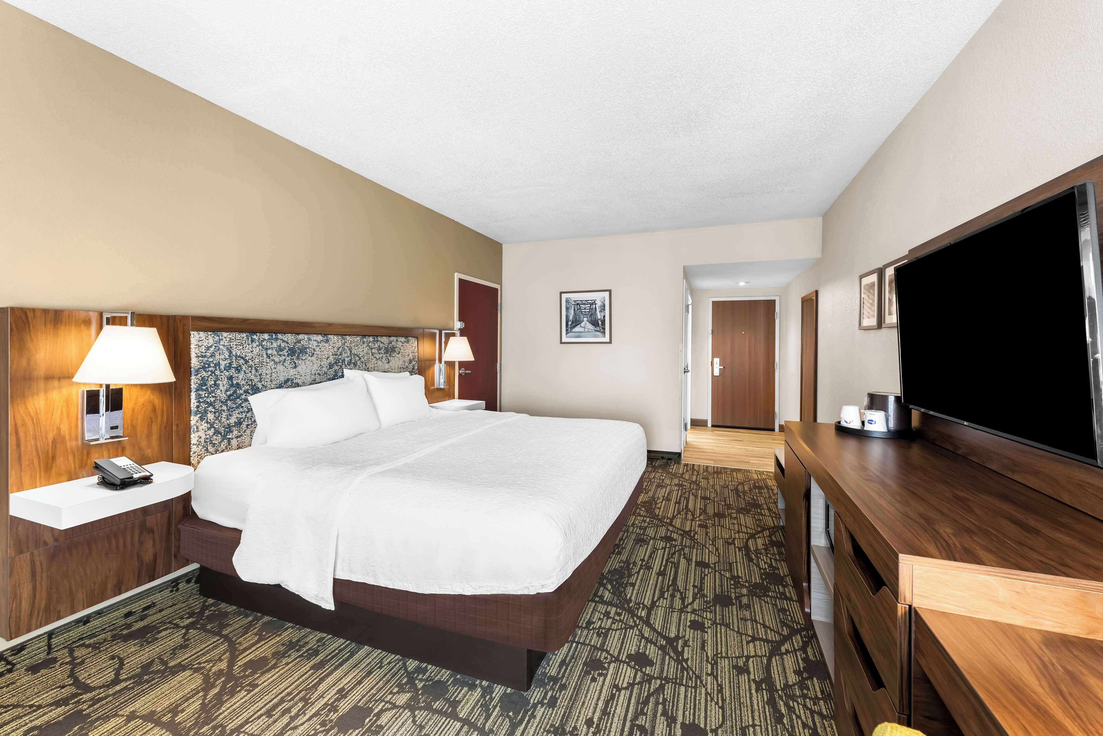 Quarto de hotel com uma cama grande, uma escrivaninha com abajur, televisão e piso coberto por carpete.