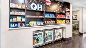 Um bar de lanches com prateleiras repletas de diversos petiscos e bebidas, um refrigerador com produtos expostos e um letreiro na parede com a palavra "OH".