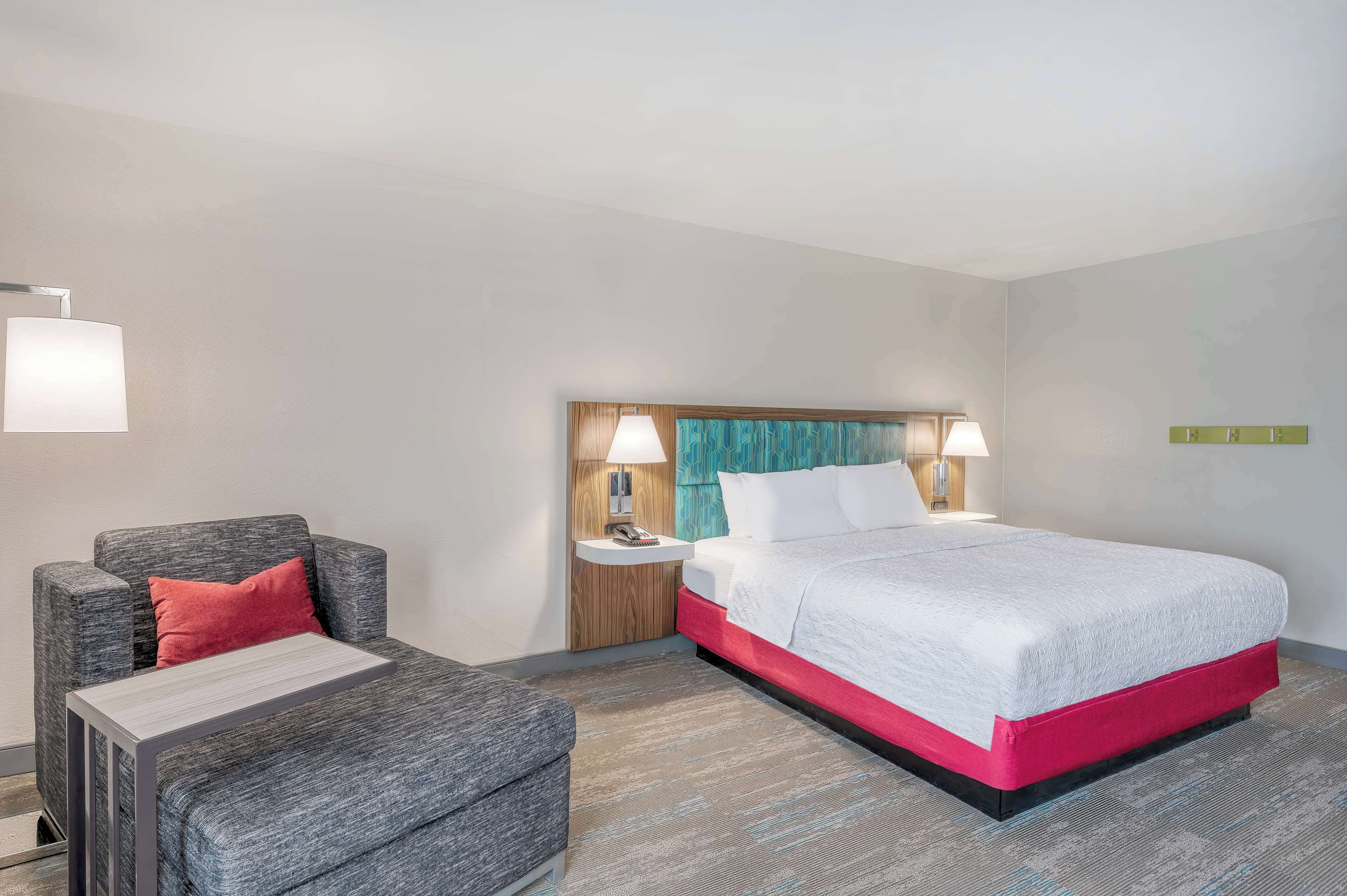 Quarto de hotel com uma cama, uma cadeira, uma mesinha de cabeceira e um abajur.
