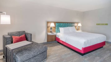 Quarto de hotel com uma cama, uma cadeira, uma mesinha de cabeceira e um abajur.