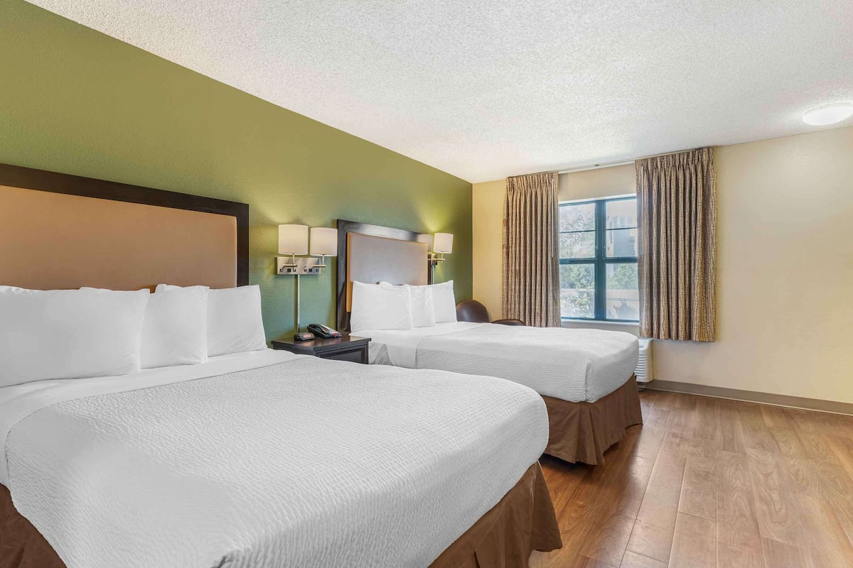 Quarto de hotel com duas camas, uma mesa de cabeceira, uma luminária e uma janela com cortinas.