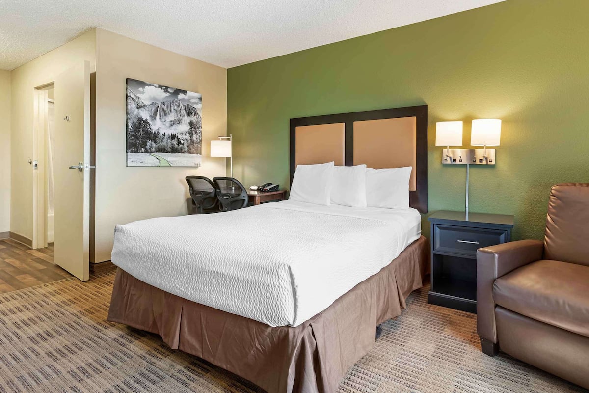 Quarto de hotel com uma cama grande, uma escrivaninha com cadeira, uma mesa de cabeceira e um abajur.
