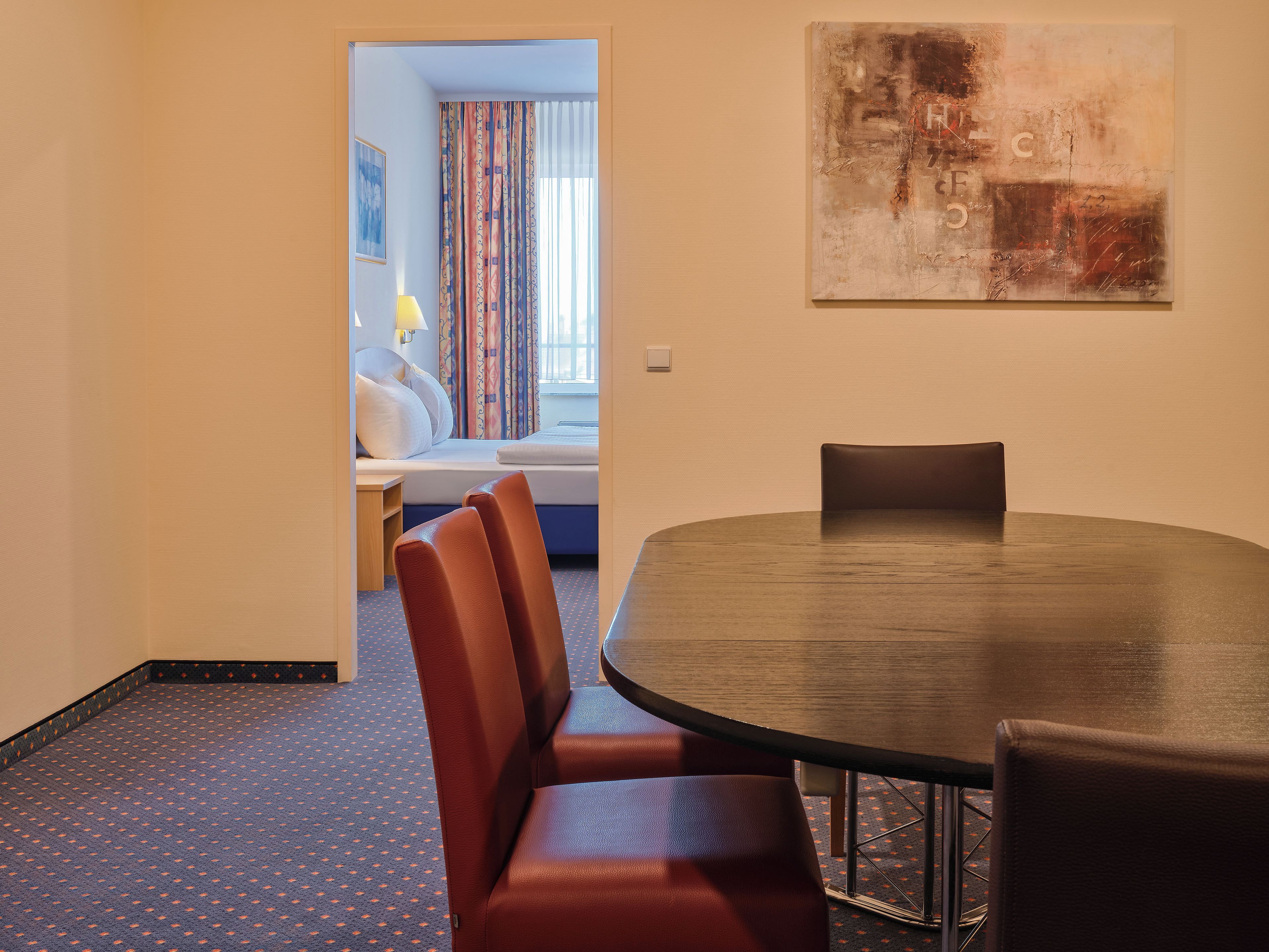 Quarto de hotel com uma mesa de jantar redonda, cadeiras estofadas em vermelho, cama com roupa de cama branca, cortina colorida e uma grande pintura abstrata na parede.