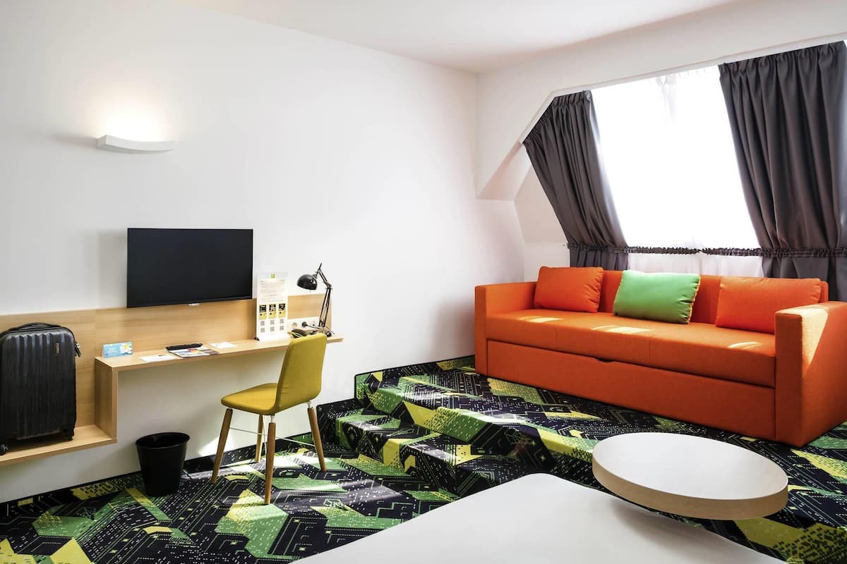 Quarto de hotel moderno com escrivaninha de madeira, TV de tela plana, sofá com almofadas, tapete estampado e mesa de centro redonda.