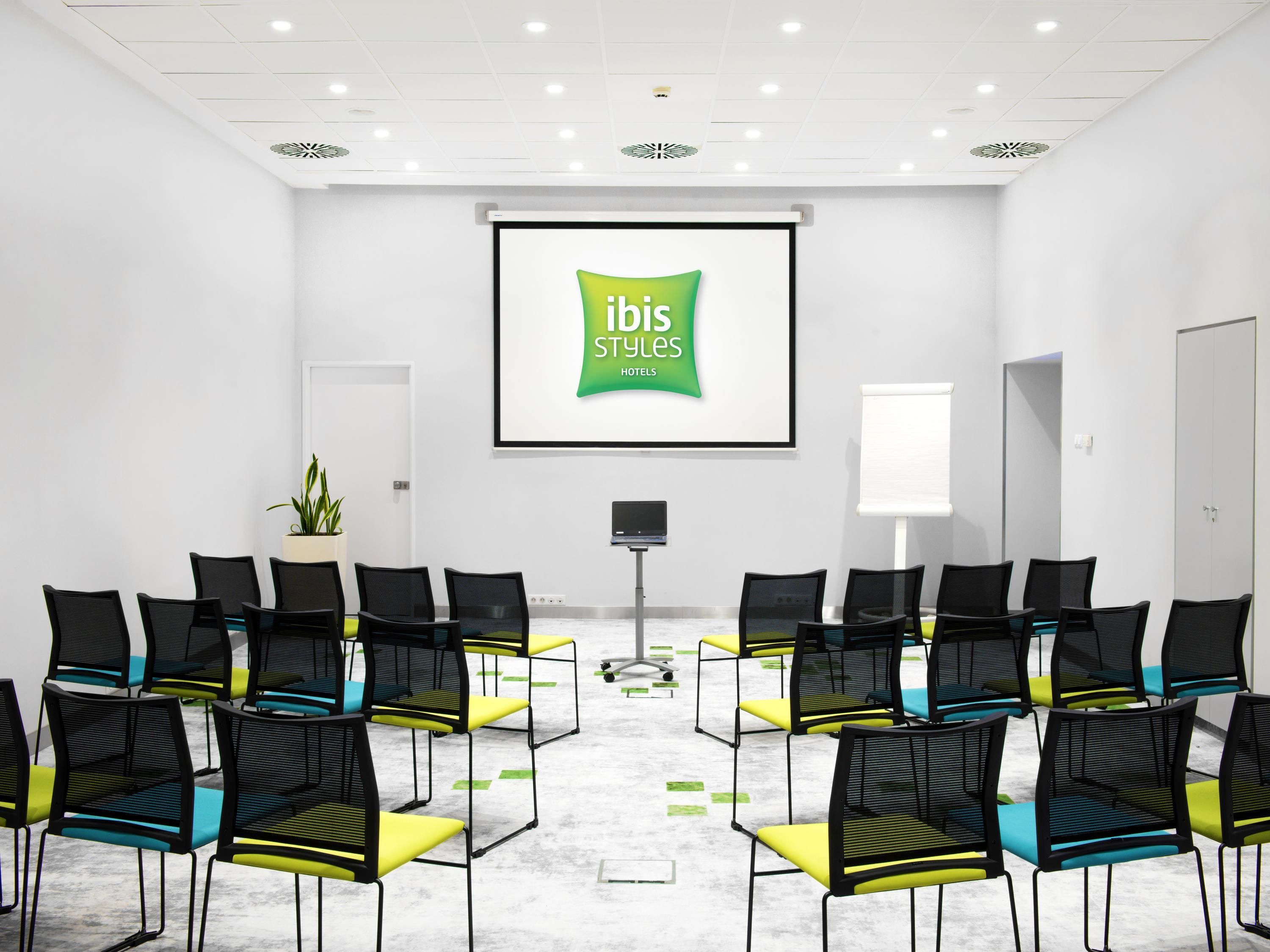 Uma sala de conferências com tela de projetor exibindo o logotipo 'ibis Styles Hotels', disposição de assentos, uma pequena mesa com um laptop e um vaso de planta.