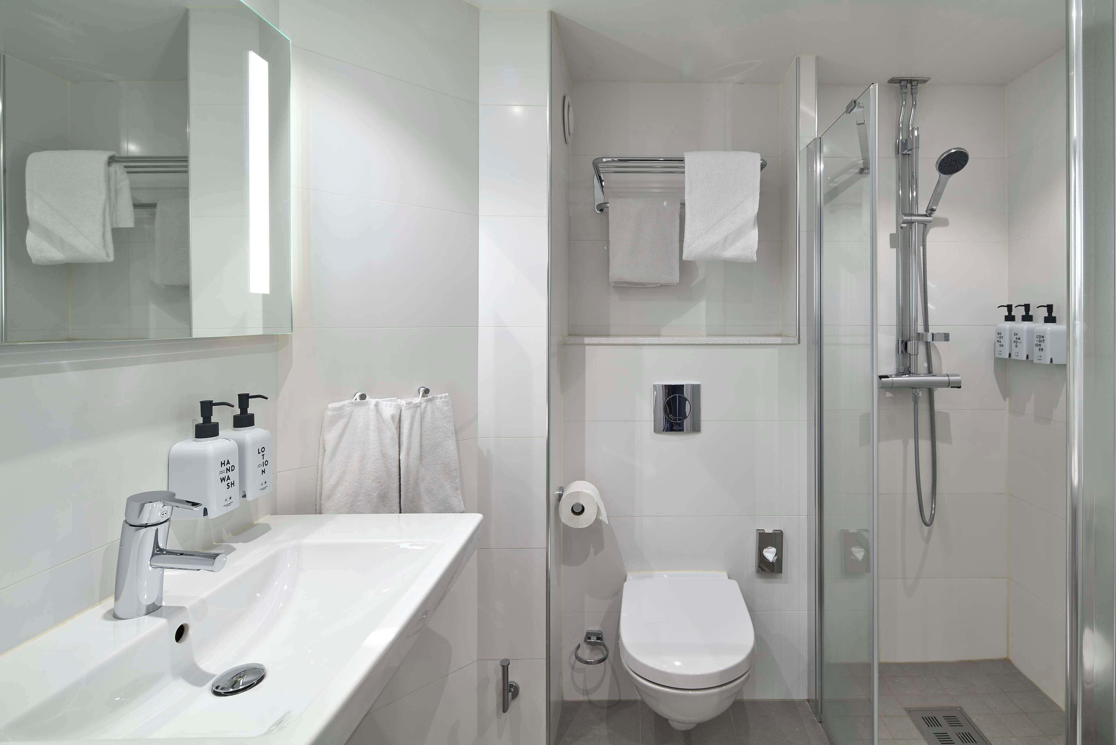 Casa de banho moderna com chuveiro envolto em vidro, lavatório branco com dispensadores de sabão e sanita com suporte para toalhas de papel.