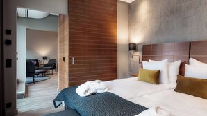 Quarto de hotel moderno com uma cama grande, um espaço de estar com uma poltrona e uma mesa pequena, e uma parede de destaque em madeira.