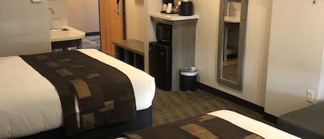 Quarto de hotel com duas camas, uma escrivaninha, máquina de café, televisão e banheiro visível por uma porta aberta.
