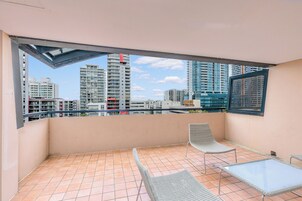 Varanda com duas cadeiras de vime e uma mesa pequena, com vista para um horizonte urbano repleto de arranha-céus.