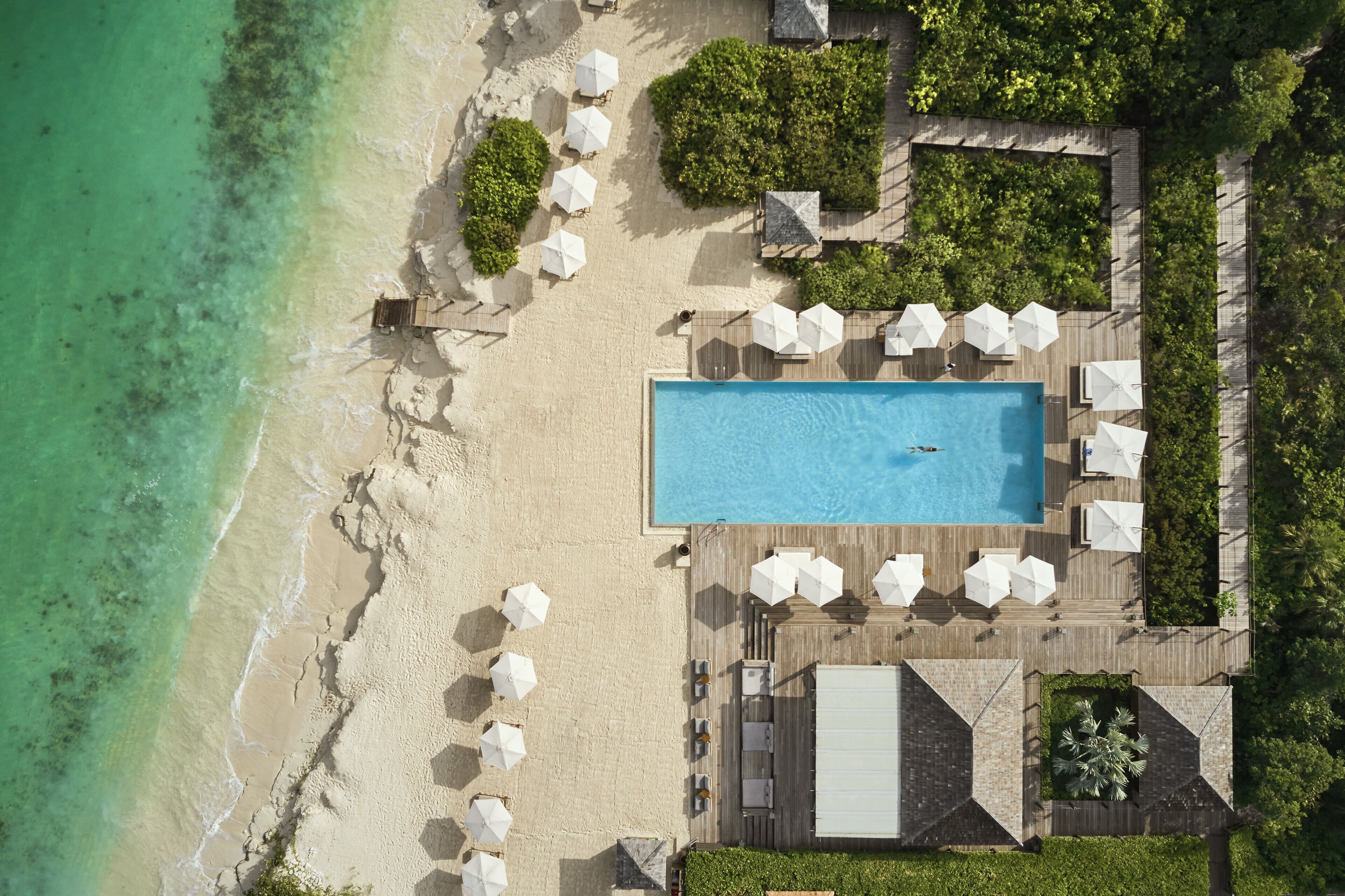 Vista aérea de um resort com piscina cercada por guarda-sóis e espreguiçadeiras, ao lado de uma praia de areia e águas turquesa cristalinas.