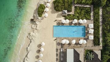 Vista aérea de um resort com piscina cercada por guarda-sóis e espreguiçadeiras, ao lado de uma praia de areia e águas turquesa cristalinas.