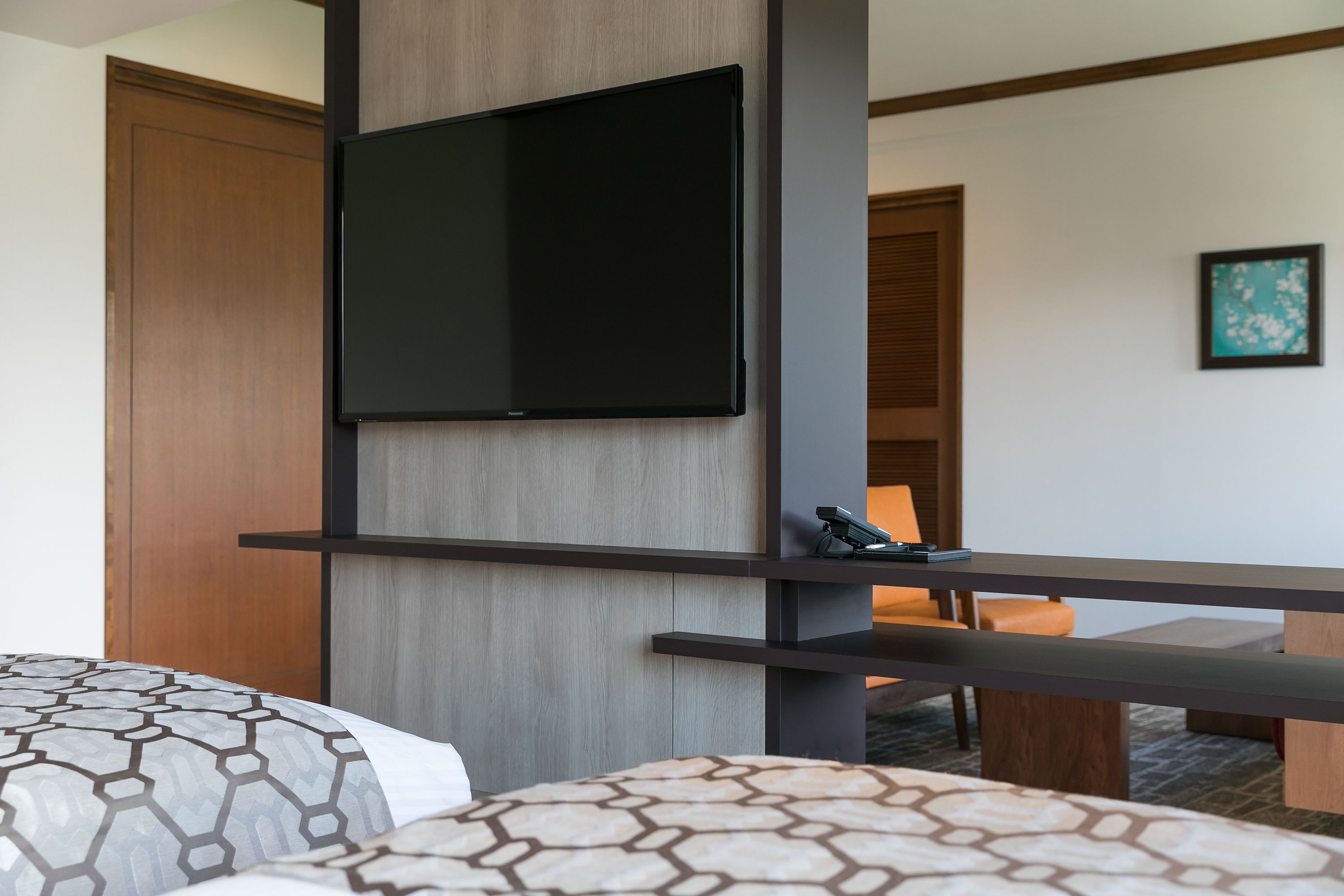Quarto de hotel com uma cama, uma televisão de tela plana presa a um painel de madeira, uma mesa com telefone e uma cadeira laranja.