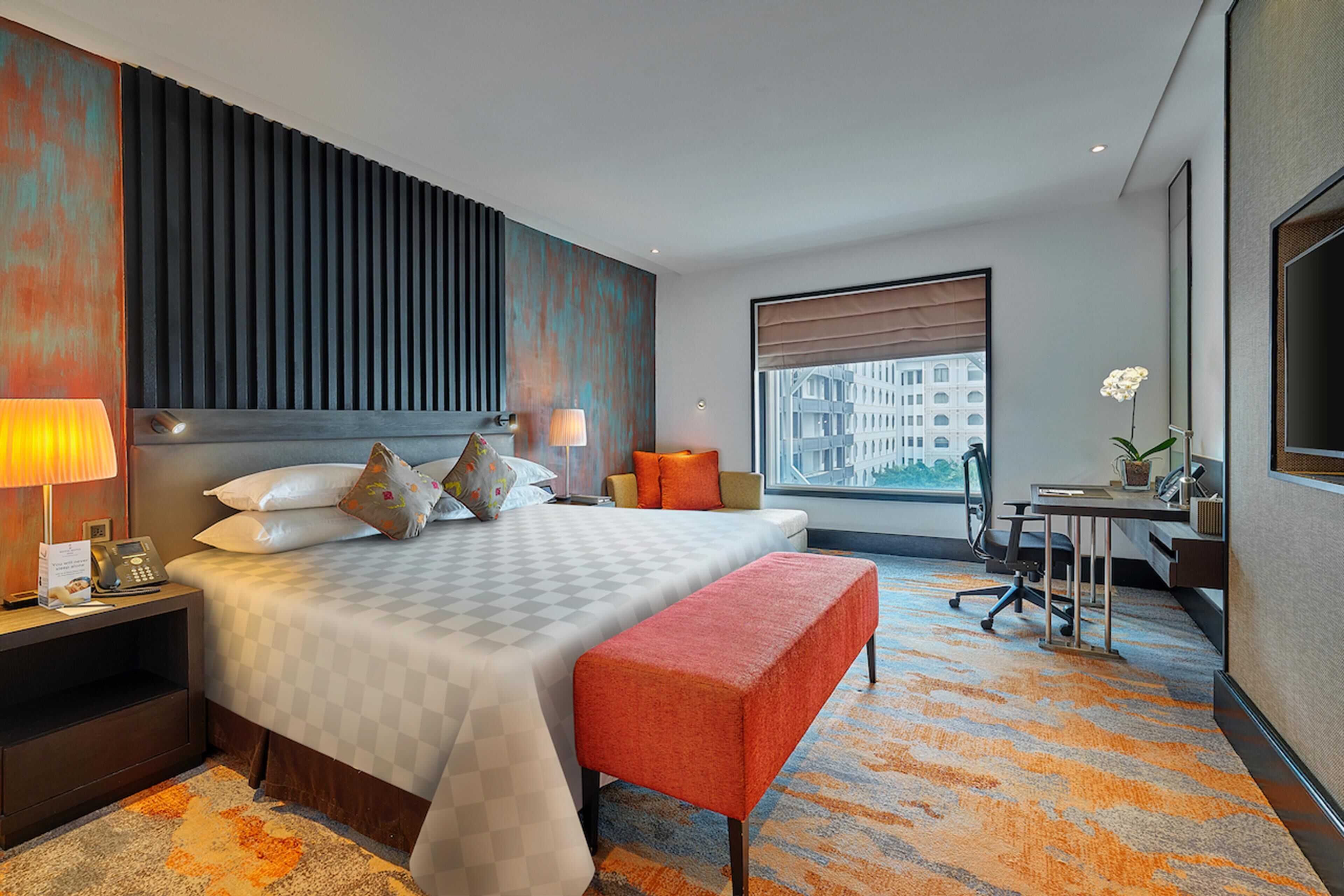 Quarto de hotel com uma cama grande, uma escrivaninha, uma cadeira, um banco, um abajur, um telefone e uma janela com vista para a cidade.