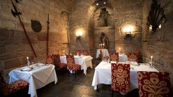 Uma sala de jantar em estilo medieval, com paredes de pedra, mesas de madeira e cadeiras vermelhas com padrão.
