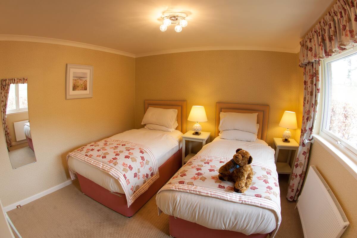 Quarto de hotel com duas camas de solteiro, cada uma com mesa de cabeceira e luminária, uma janela com cortinas, um radiador e um quadro na parede.