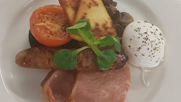 Um prato com uma fatia de presunto, ovo pochê, cogumelos grelhados, batatas assadas e uma fatia de tomate.
