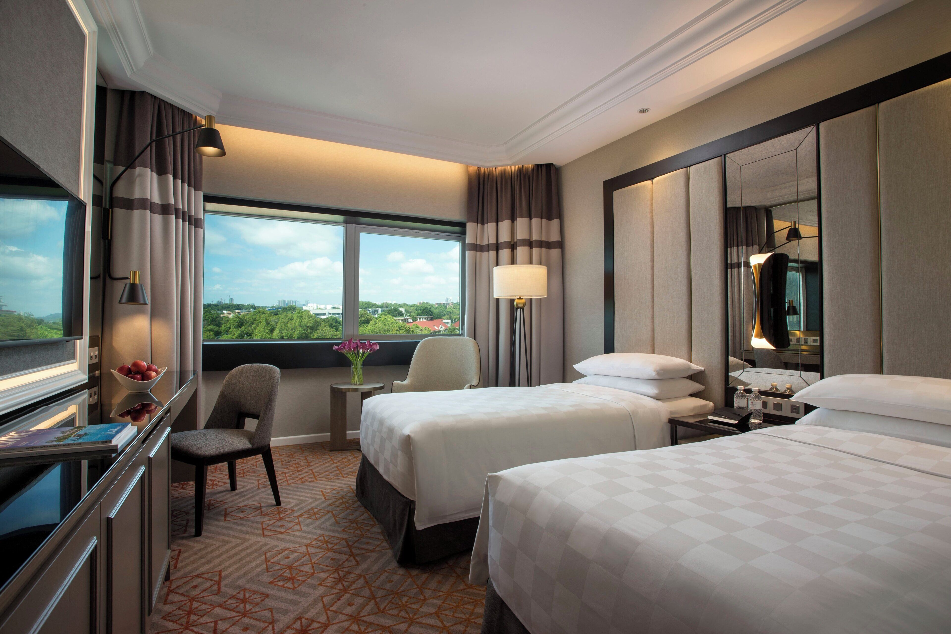 Quarto de hotel com duas camas, uma escrivaninha com computador, uma cadeira, uma mesinha com um vaso de flores e uma janela grande com vista para a cidade.