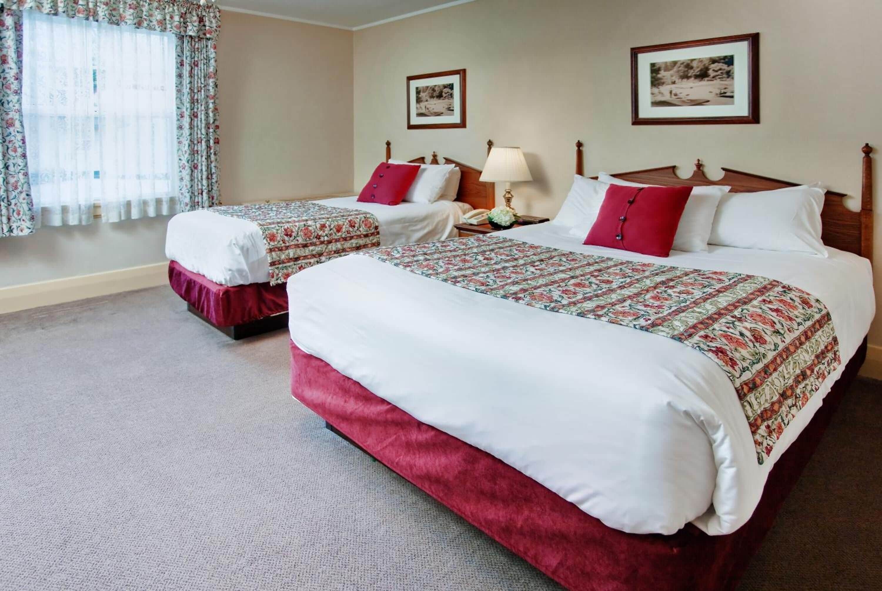 Quarto de hotel com duas camas, cada uma com colcha vermelha e branca, cabeceira de madeira e mesa de cabeceira com abajur.