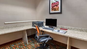 Quarto de hotel com escrivaninha, computador, cadeira e um quadro na parede.