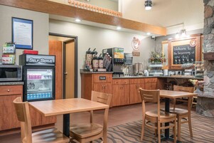 Uma estação de café autoatendimento com geladeira, micro-ondas e máquinas de café. Há mesas e cadeiras de madeira, e um quadro negro com uma mensagem de boas-vindas aos hóspedes.