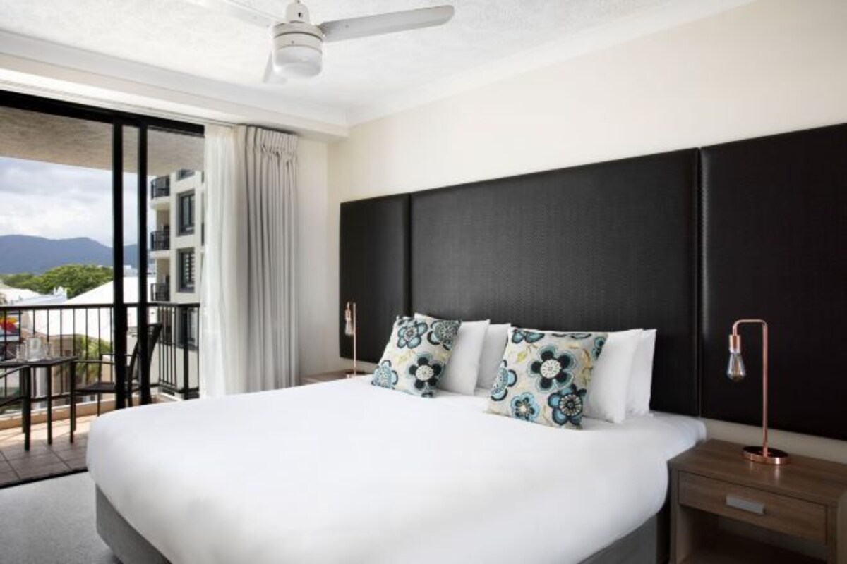 Quarto de hotel com uma cama grande, um ventilador de teto, uma varanda com vista para edifícios e montanhas, e uma mesa de cabeceira com uma luminária.