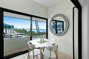 Uma varanda com mesa e cadeiras, uma janela grande com vista para o mar e edifícios, e um espelho decorativo na parede.