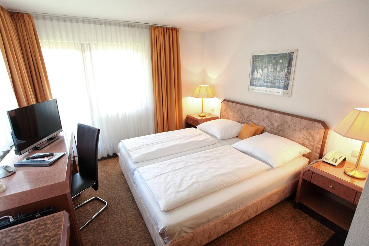 Quarto de hotel com cama, escrivaninha, televisão, cadeira e mesa de cabeceira com luminária.