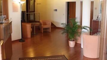 Uma área de recepção de hotel com um tapete castanho, balcão de recepção, planta em vaso e um espaço de estar com cadeiras.