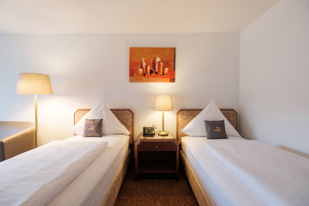 Quarto de hotel com duas camas de solteiro, cada uma com mesa de cabeceira, abajur e almofada decorativa.