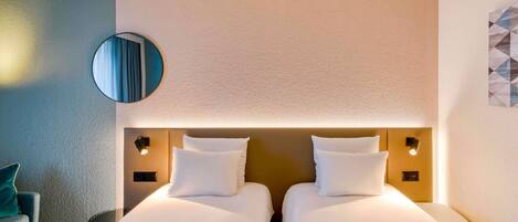 Quarto de hotel com duas camas, um espelho redondo, abajures, uma mesinha e uma cadeira.
