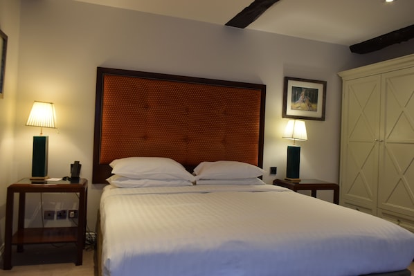 Uma cama bem arrumada com lençóis brancos, duas mesas de cabeceira com abajures, uma cabeceira com padrão texturizado e um guarda-roupa com design de cruz.