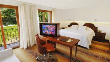 Quarto de hotel com cama, escrivaninha, televisão, cadeira e varanda com cortinas.