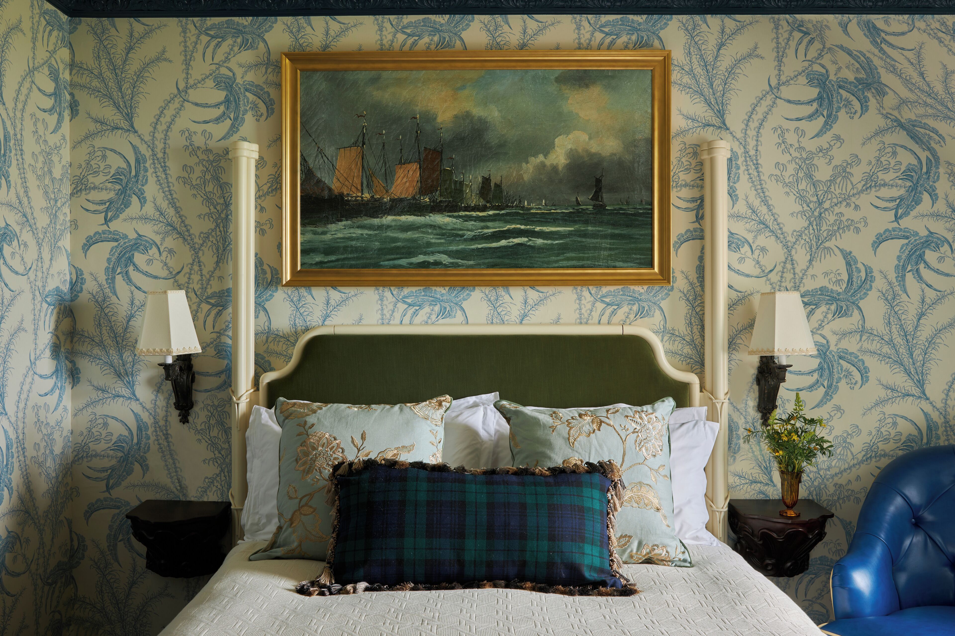 Uma cama com cabeceira verde, roupa de cama branca e almofada xadrez. Há duas mesas de cabeceira com luminárias e um quadro de um navio na parede.
