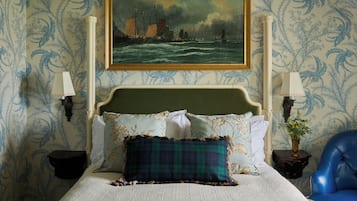 Uma cama com cabeceira verde, roupa de cama branca e almofada xadrez. Há duas mesas de cabeceira com luminárias e um quadro de um navio na parede.