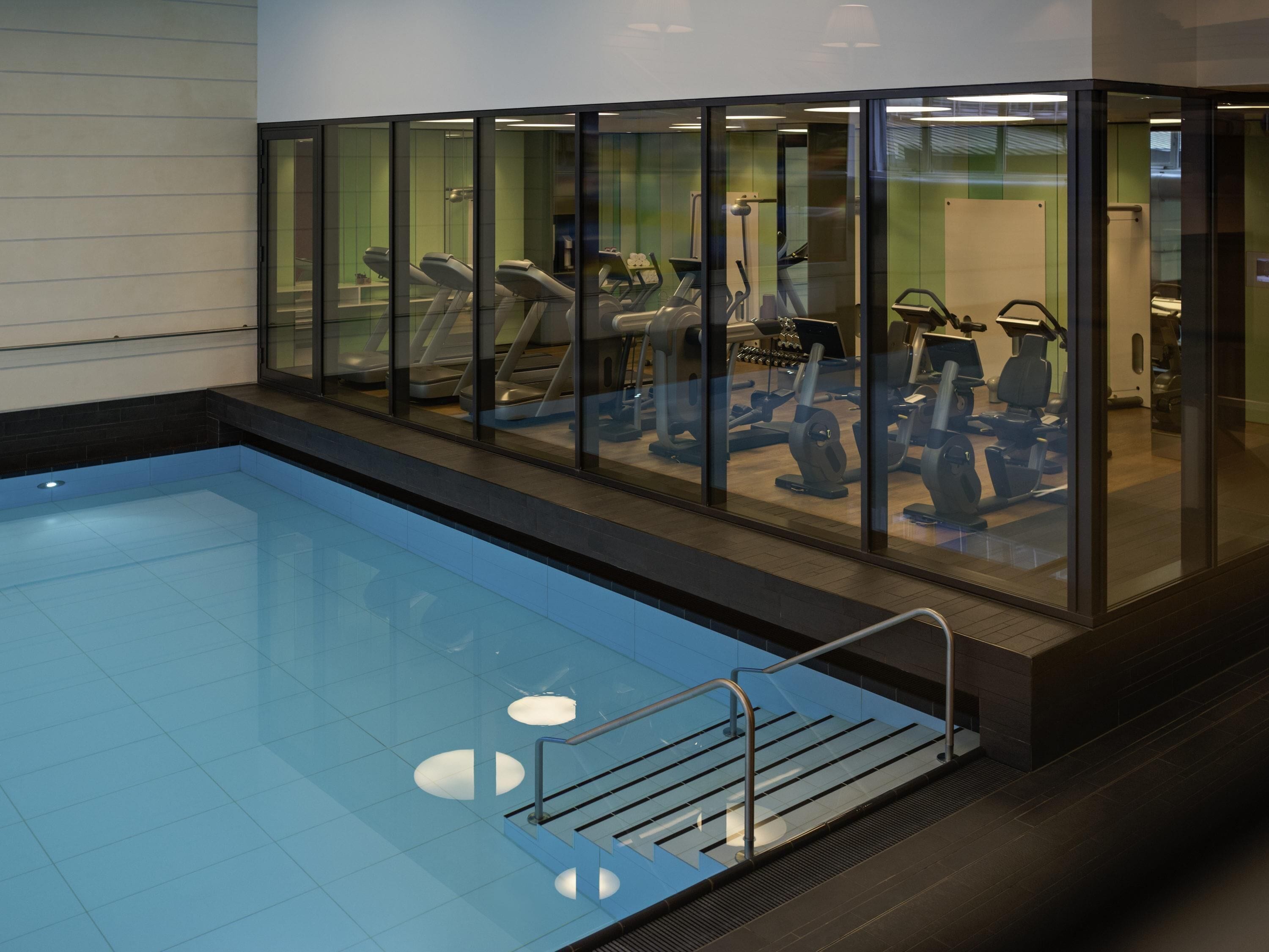 Piscina interior moderna com cobertura de vidro, cercada por equipamentos de ginástica.