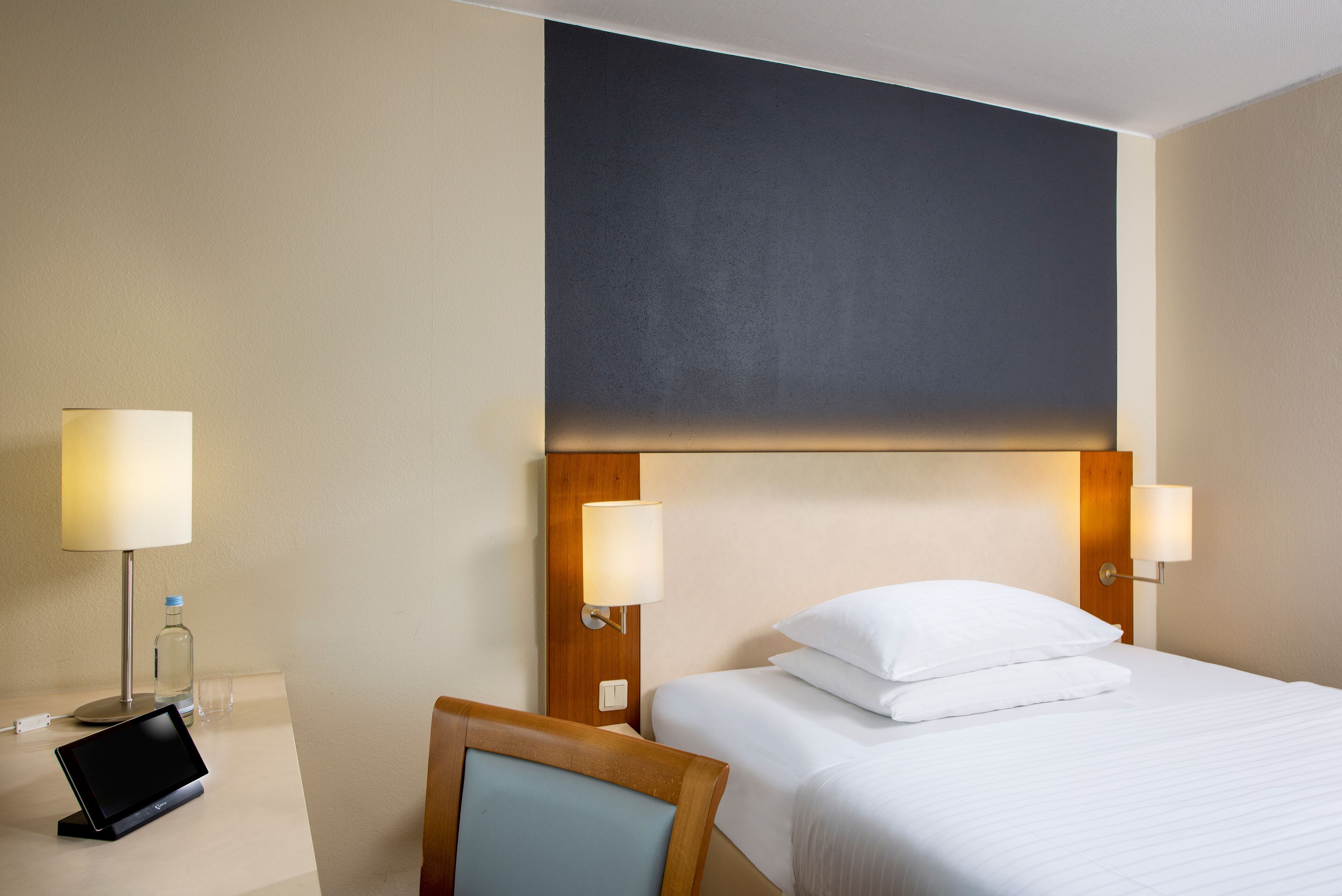 Quarto de hotel com cama, luminárias de cabeceira, mesa de cabeceira com abajur, uma cadeira e um tablet.
