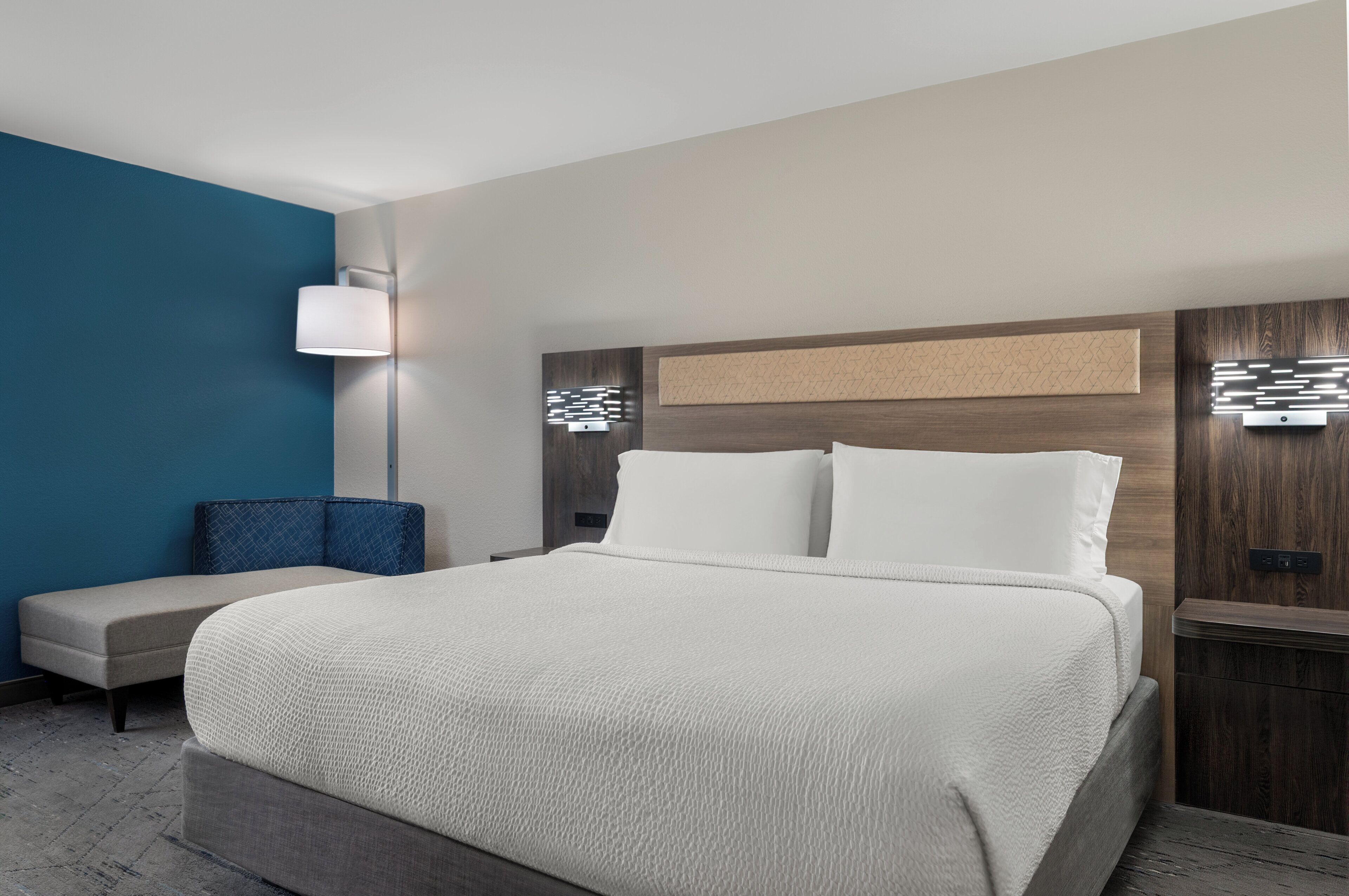 Quarto de hotel moderno com uma cama grande, uma mesinha de cabeceira, uma luminária e uma parede azul de destaque.