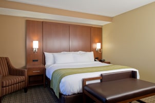 Quarto de hotel com uma cama grande, cabeceira de madeira, duas luminárias de cabeceira, uma mesa de cabeceira, uma poltrona listrada e um banco marrom.