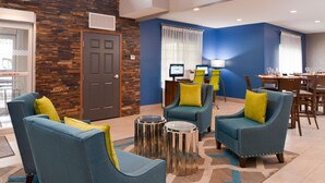 Um hall de hotel com poltronas azuis e amarelas, uma parede de destaque em madeira e uma área de refeições com mesas e cadeiras.