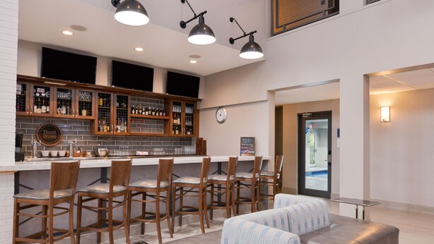 Uma área de bar moderna com cadeiras de madeira, balcão e um espaço de estar com sofá e apoio para os pés.