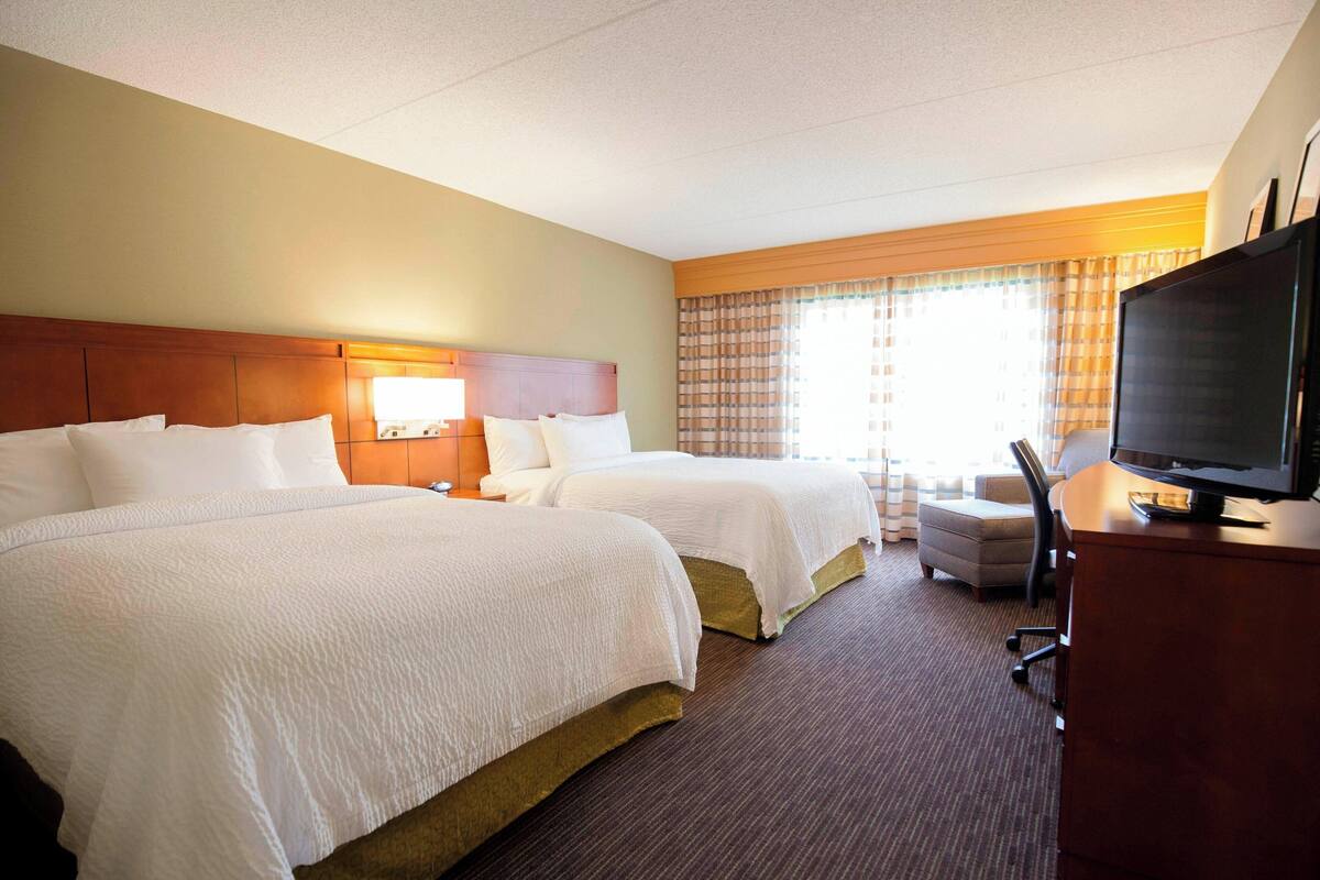 Quarto de hotel com duas camas, uma escrivaninha, televisão e janela com cortinas.