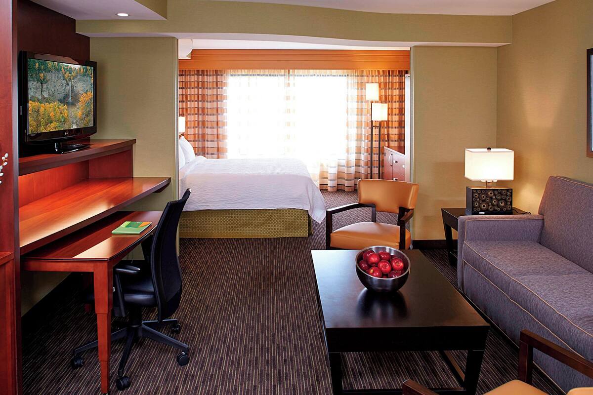 Quarto de hotel com cama, escrivaninha com computador, televisão, sofá, mesa de centro com frutas e abajur.