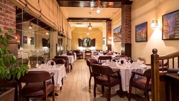Restaurante com várias mesas preparadas para refeições, com paredes de tijolo e vigas de madeira.