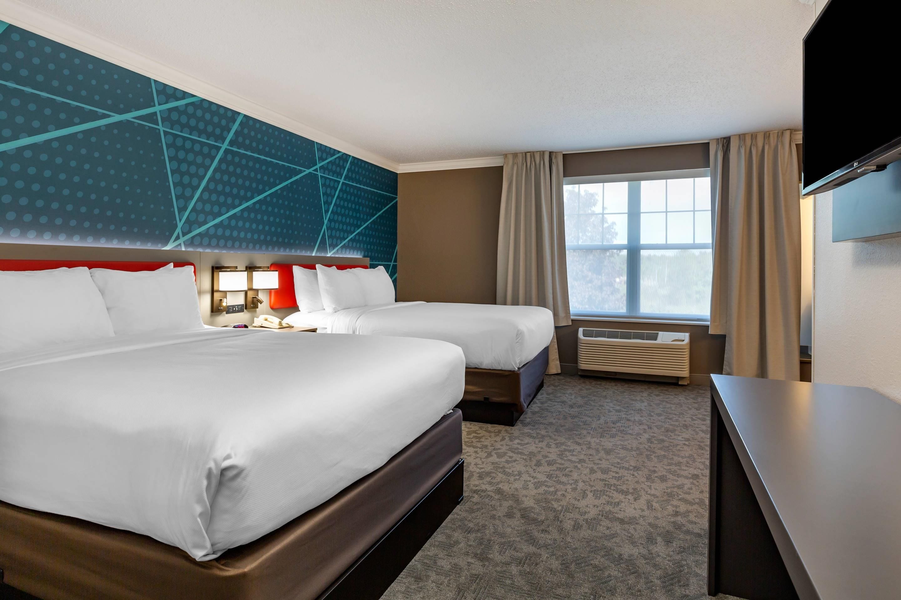 Quarto de hotel com duas camas, uma TV de tela plana, janela com cortinas e ar-condicionado fixado na parede.