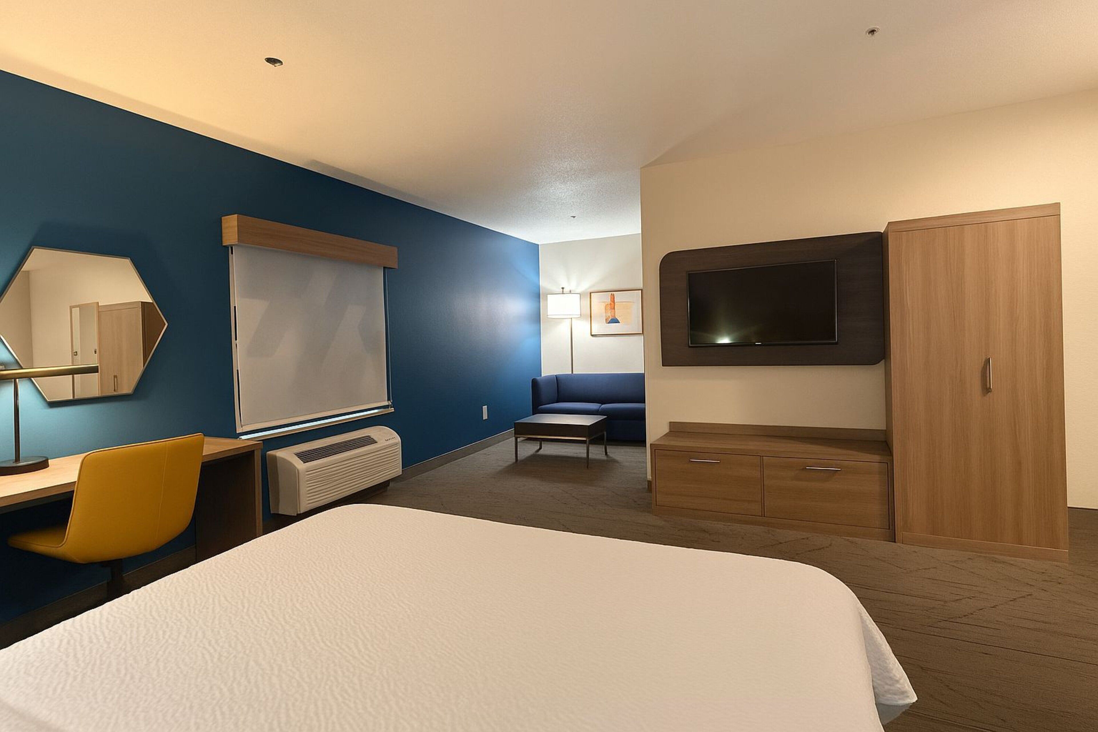 Quarto de hotel com uma cama, uma escrivaninha com cadeira, um sofá, uma TV fixada na parede e um guarda-roupa.
