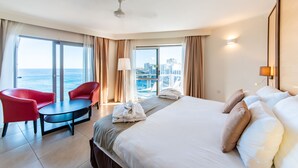 Quarto de hotel com uma cama, uma mesa, duas cadeiras vermelhas, vista para o oceano e para a paisagem urbana através de janelas amplas.