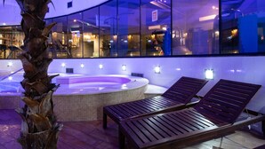 Uma área de piscina interior moderna com uma grande janela, uma palmeira e espreguiçadeiras de madeira.